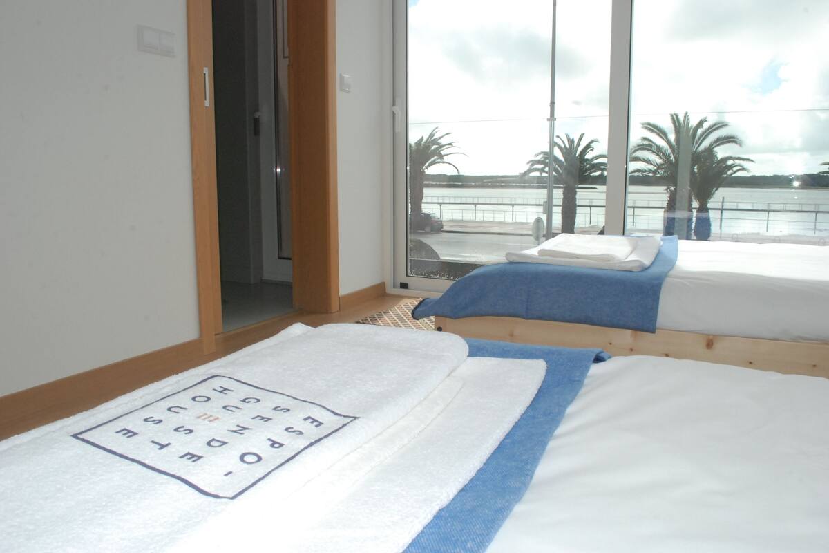 Quarto de hotel com uma grande janela com vista para um corpo de água e palmeiras. Há duas camas com toalhas, uma com toalha azul e outra com toalha branca.