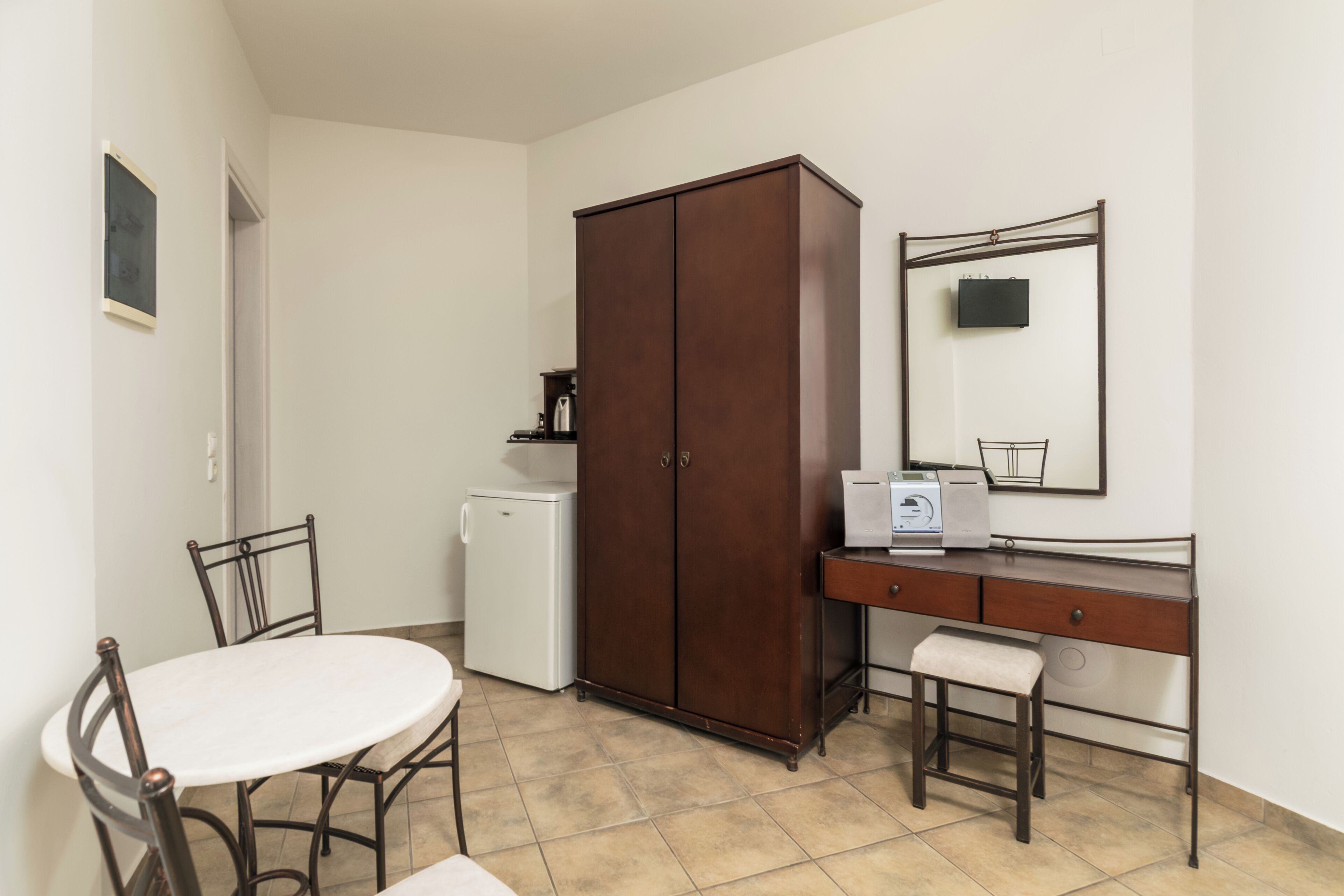 Quarto pequeno com mesa de jantar, cadeiras, guarda-roupa, mini-geladeira, escrivaninha com cadeira e espelho.