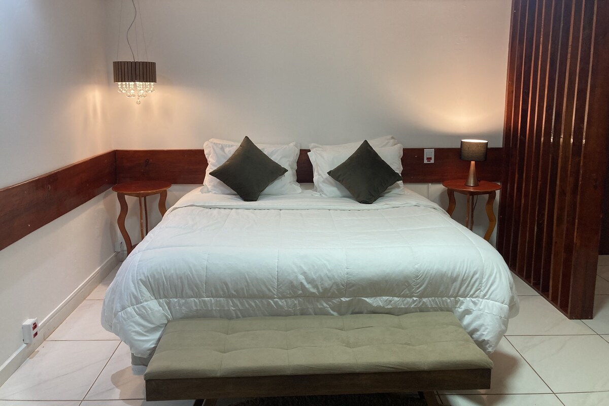 Uma cama bem arrumada com cabeceira, duas mesas de cabeceira, um banco e um lustre.