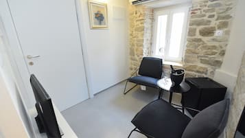 Quarto pequeno e acolhedor com parede de pedra, porta branca, cadeira preta, mesa com um copo e um alto-falante preto.