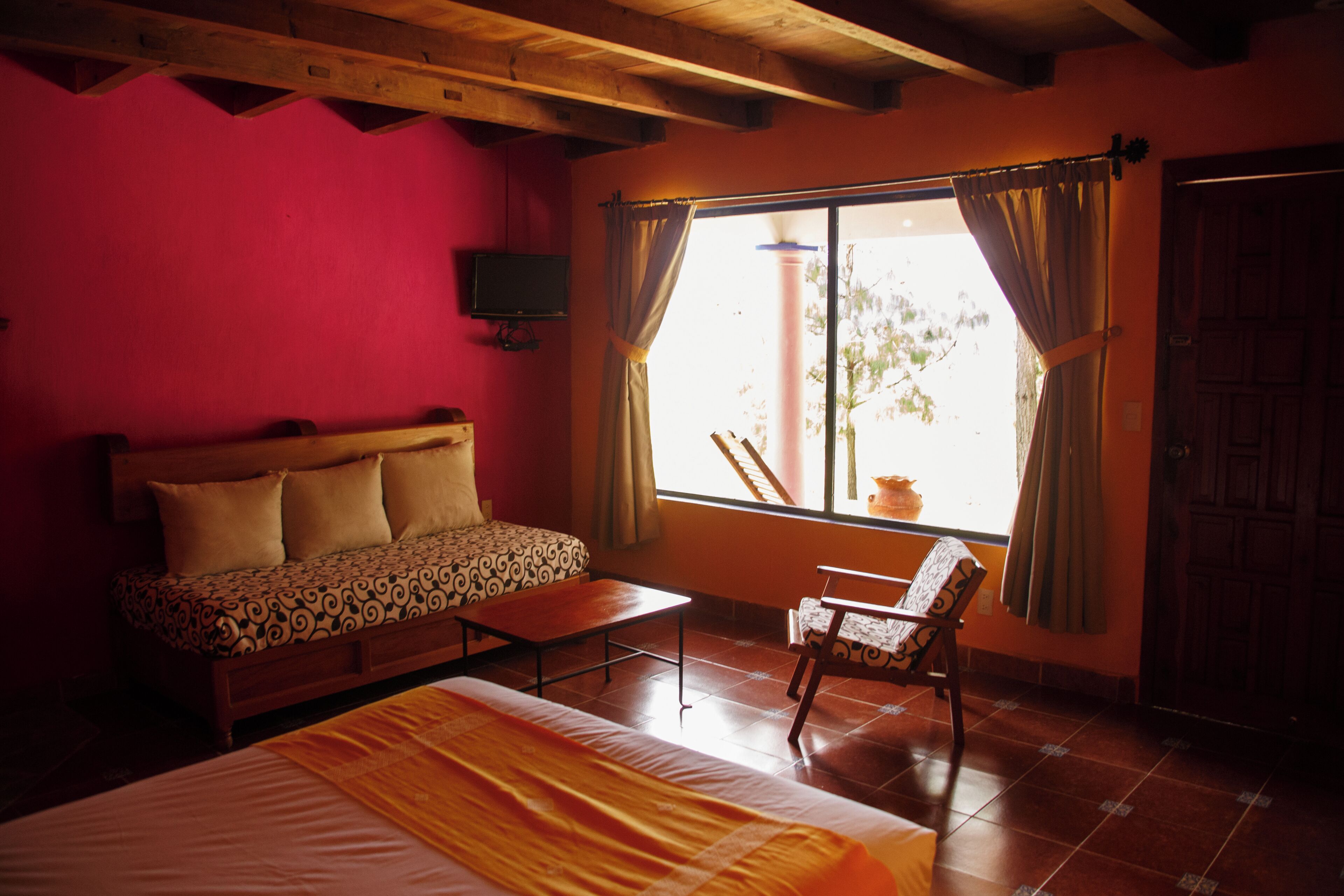 Um quarto com cama, sofá, poltrona, mesa pequena, televisão e janela com cortinas.