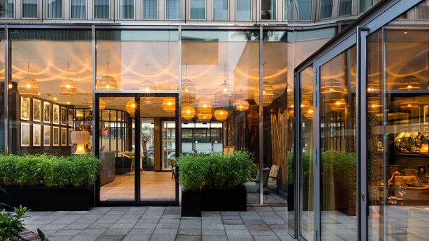 Entrada de hotel moderna com portas de vidro, plantas internas e luminárias pendentes.