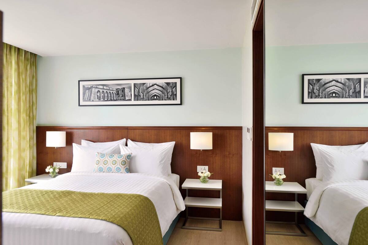 Quarto de hotel com duas camas, cabeceira de madeira, mesas de cabeceira com luminárias e quadros na parede.