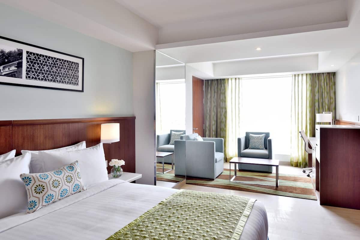 Quarto de hotel com uma cama grande, uma escrivaninha, uma cadeira e um sofá. Há uma janela com cortinas e vista para o exterior.
