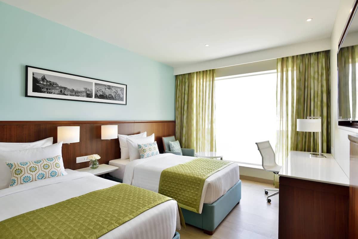 Quarto de hotel com duas camas, uma escrivaninha e uma cadeira. Há uma janela com cortinas e um quadro na parede.