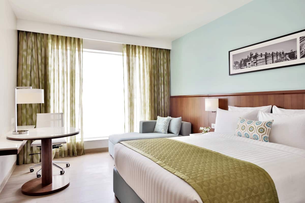 Quarto de hotel com cama, escrivaninha, cadeira e janela com cortinas.