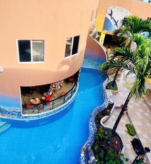 Uma área de piscina com borda curva, uma área de refeições com mesas e cadeiras, e uma variedade de plantas e arbustos.