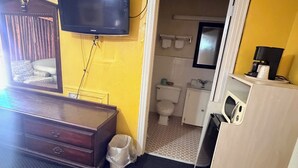 Quarto de hotel com cômoda de madeira, televisão fixada na parede, uma pequena lixeira e cafeteira.