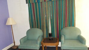 Um quarto com duas poltronas azul-turquesa, uma pequena mesa de madeira, um abajur e uma cortina com padrão paisley.