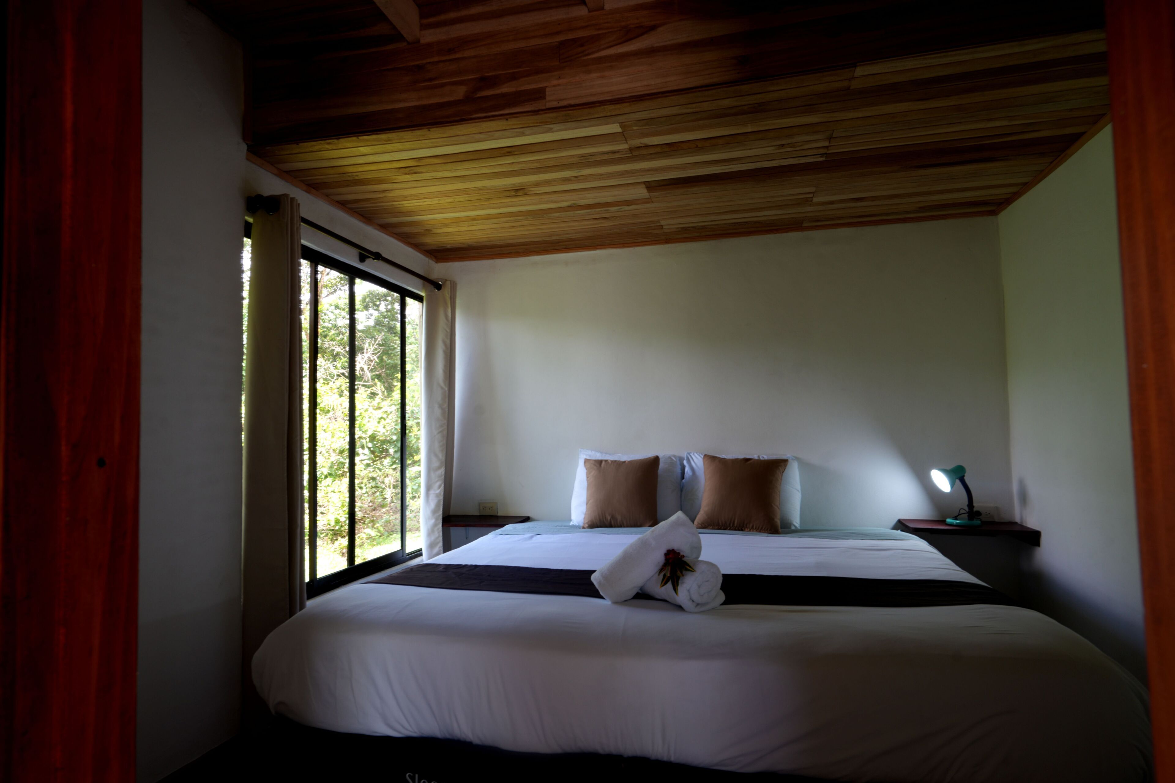 Um quarto com uma cama grande, uma mesa de cabeceira com abajur e uma janela com cortinas.