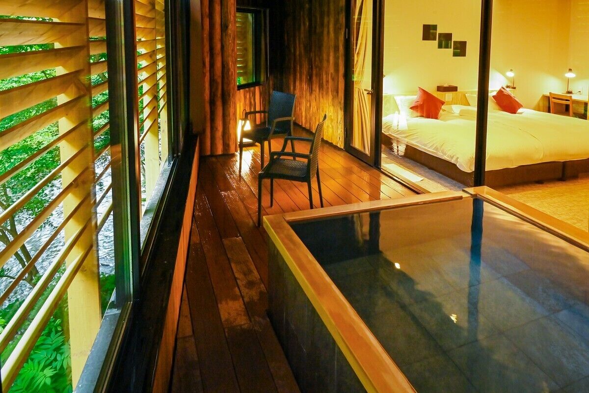 Quarto de hotel moderno com varanda envidraçada, piso de madeira e cama com roupa de cama branca e almofadas vermelhas.