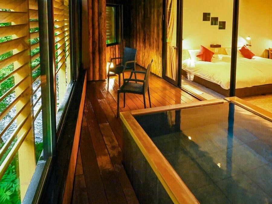 Quarto de hotel moderno com varanda envidraçada, piso de madeira e cama com roupa de cama branca e almofadas vermelhas.