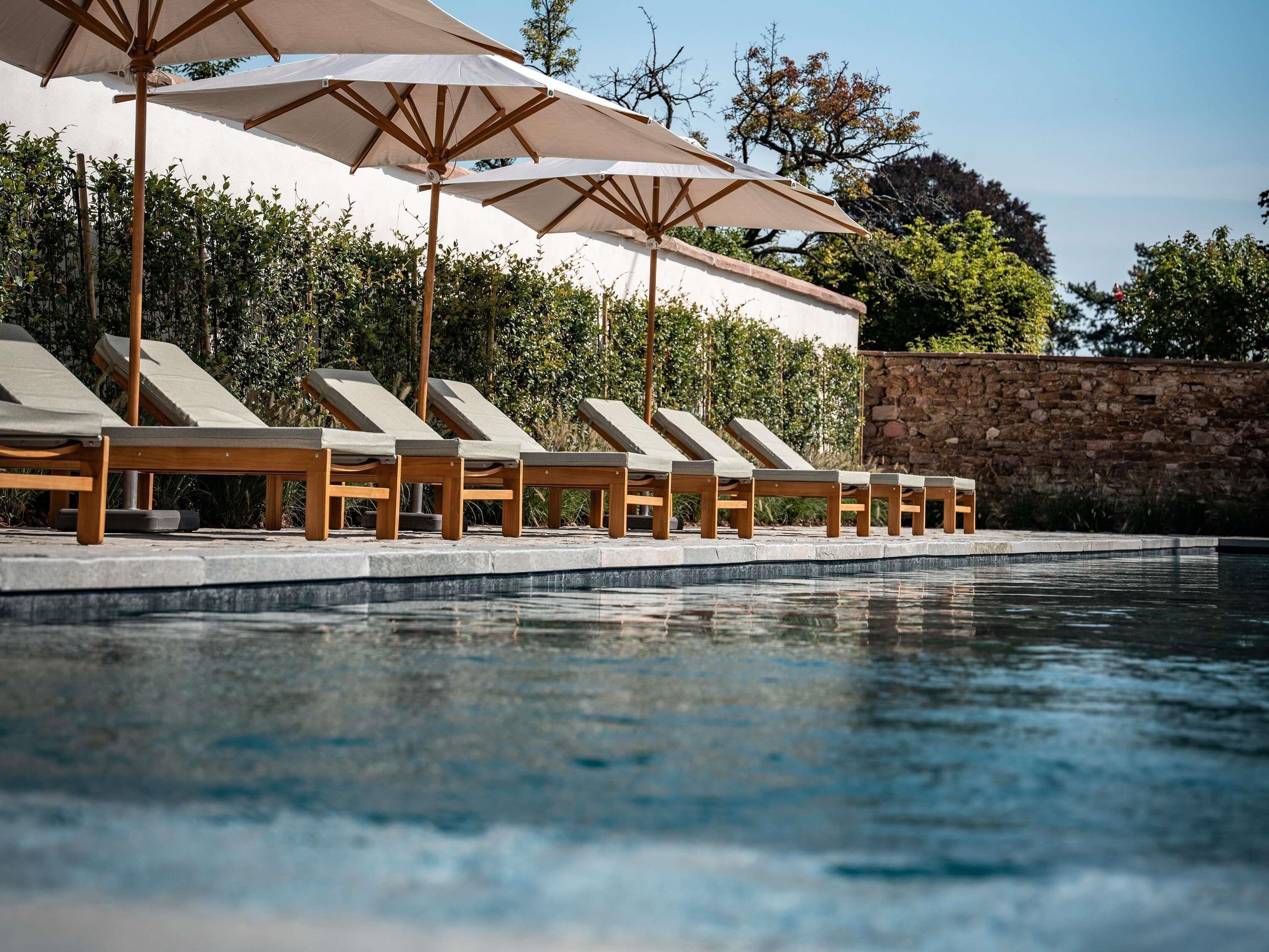Uma área na piscina com espreguiçadeiras e guarda-sóis, uma parede de pedra e vegetação ao fundo.