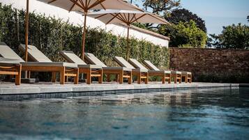 Uma área na piscina com espreguiçadeiras e guarda-sóis, uma parede de pedra e vegetação ao fundo.