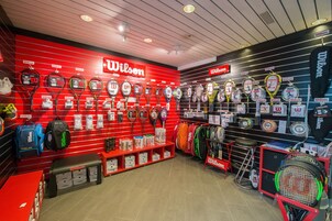Loja de artigos esportivos com paredes vermelhas, diversos raquetes de tênis e sacolas expostas em prateleiras e suportes.