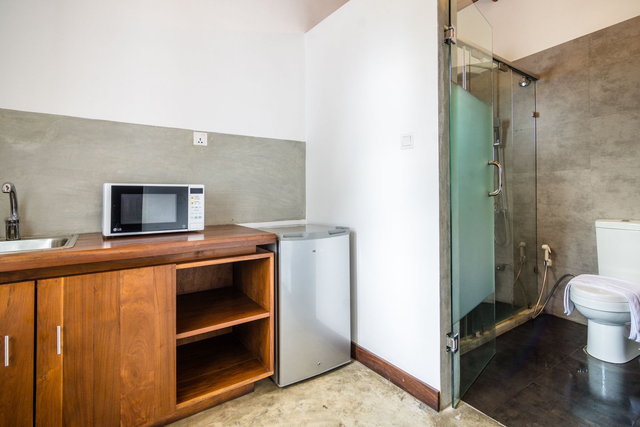Uma casa de banho compacta com toucador de madeira, micro-ondas, frigorífico pequeno, cabine de duche em vidro e sanita.