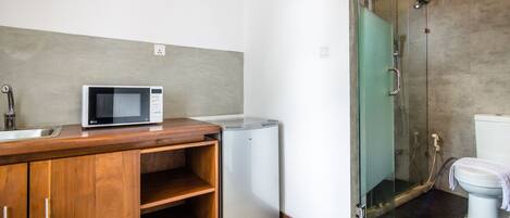 Uma casa de banho compacta com toucador de madeira, micro-ondas, frigorífico pequeno, cabine de duche em vidro e sanita.