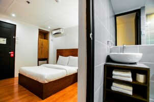 Quarto de hotel com cama de madeira, parede branca e banheiro com pia e porta-toalhas.