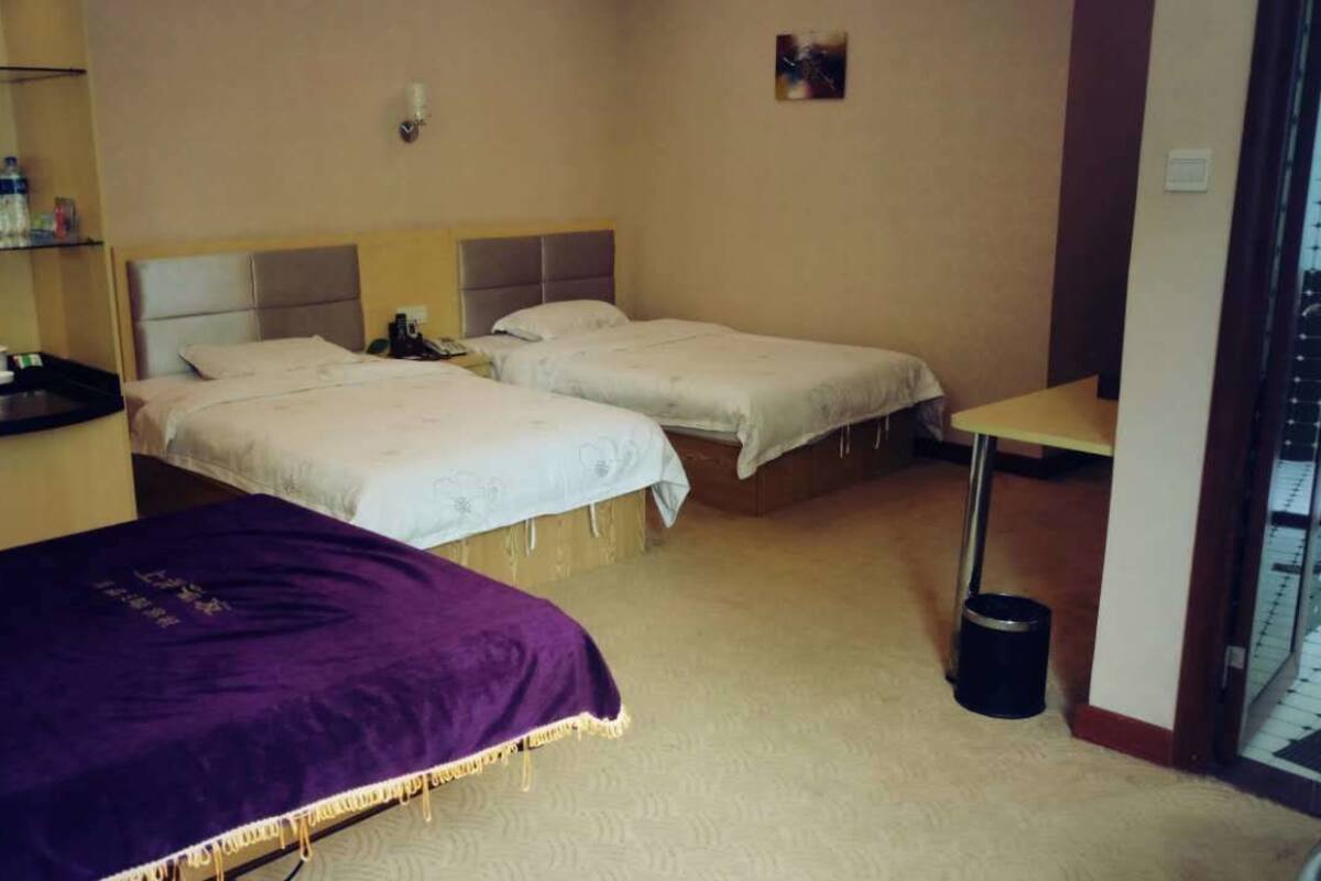 Quarto de hotel com duas camas de solteiro, uma mesa pequena, uma cadeira, prateleira com artigos de higiene pessoal e uma lixeira.