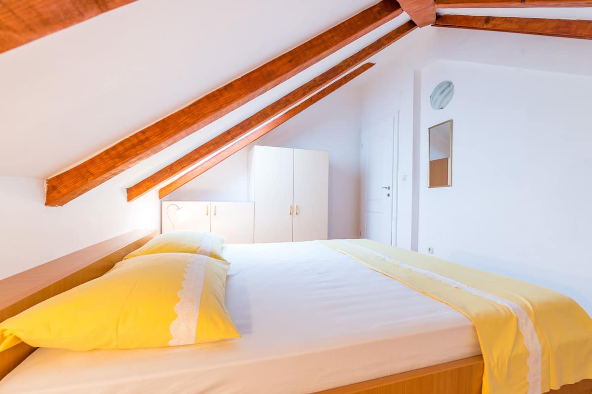 Quarto com teto inclinado, cama com roupa de cama branca e almofadas amarelas, guarda-roupa branco e um pequeno espelho na parede.