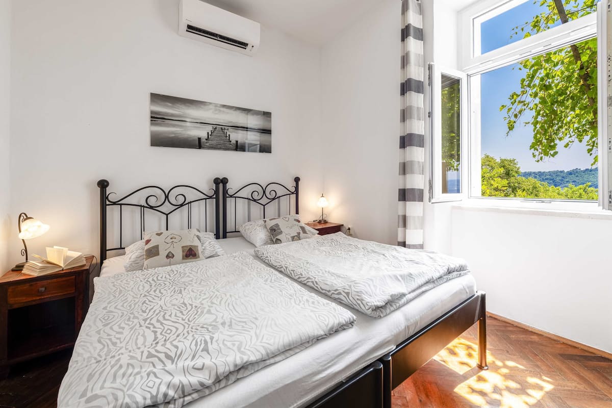 Quarto com cama, mesas de cabeceira, ar-condicionado fixado na parede, um quadro emoldurado e janela com vista para o verde.