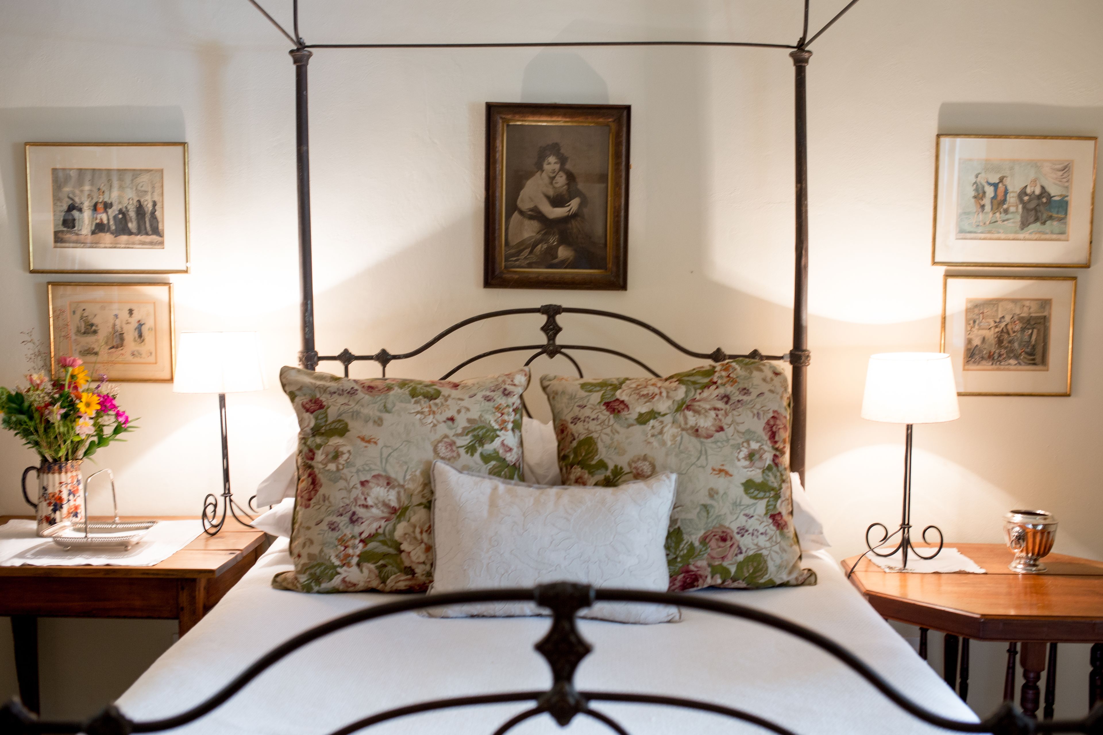 Quarto bem arrumado com cama de dossel, almofadas estampadas com motivos florais e quadros nas paredes.