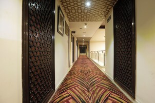 Corredor de hotel com carpete estampado, portas de madeira e sinal de elevador.