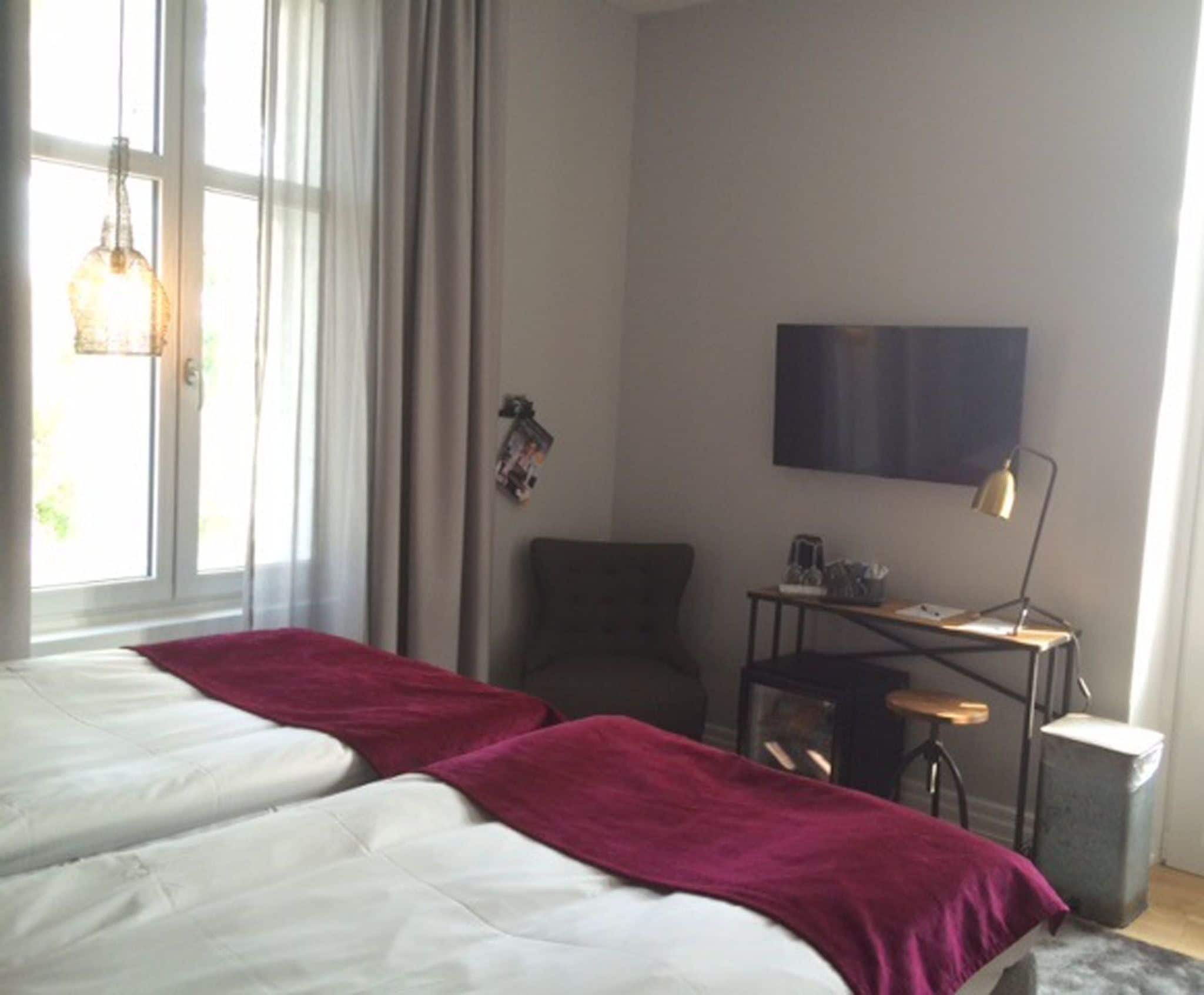 Quarto de hotel com duas camas, uma escrivaninha com cadeira, televisão na parede e janela com cortinas.