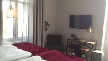 Quarto de hotel com duas camas, uma escrivaninha com cadeira, televisão na parede e janela com cortinas.