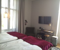 Quarto de hotel com duas camas, uma escrivaninha com cadeira, televisão na parede e janela com cortinas.
