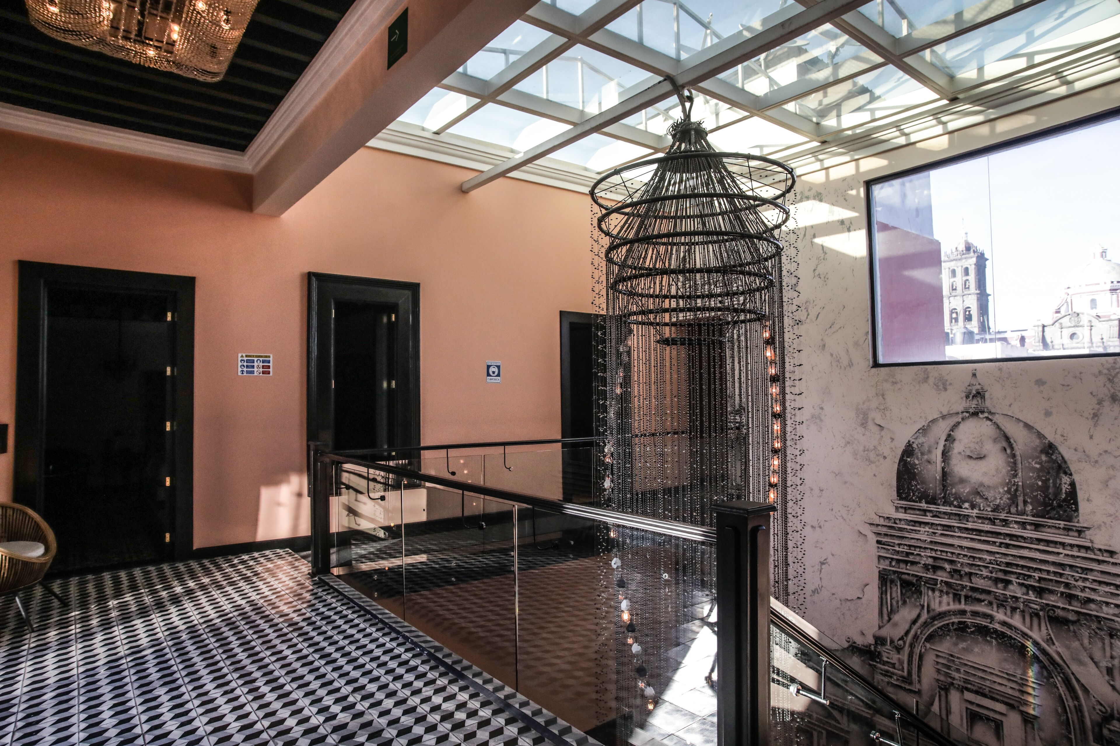 Um saguão de hotel com balcão de recepção, um candelabro, uma escadaria e um mural de um edifício.