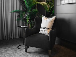 Uma poltrona preta com uma almofada decorativa, uma mesinha redonda pequena e uma planta em vaso em um quarto moderno com cortinas cinzas e uma janela.