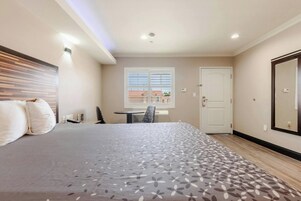 Quarto de hotel com cama, escrivaninha, cadeira, janela com persianas e porta.