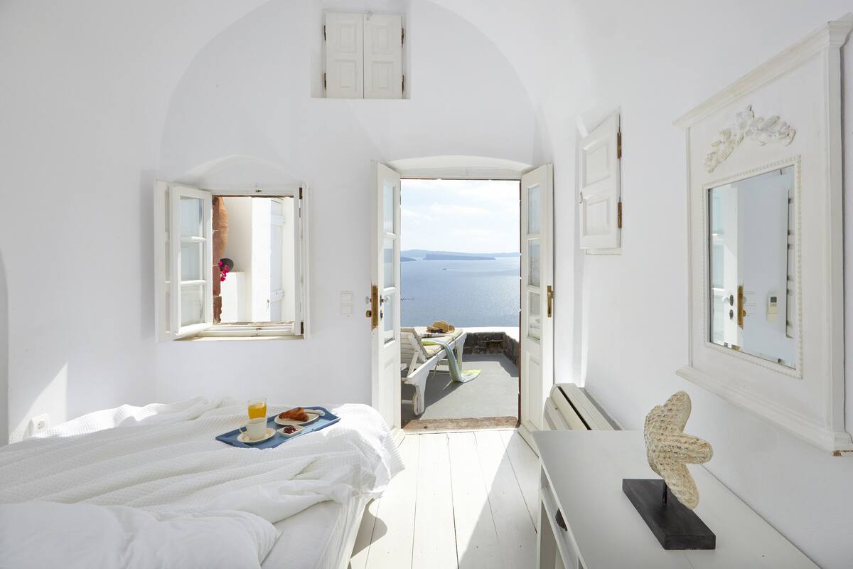 Um quarto claro e branco com uma cama, uma mesa com uma escultura, e vista para o mar através de uma porta aberta.
