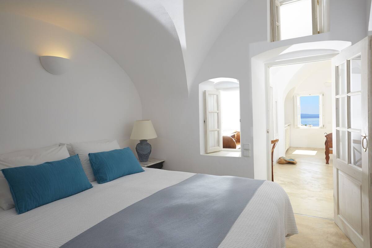 Quarto com cama, travesseiros, abajur e porta aberta para uma varanda com vista.