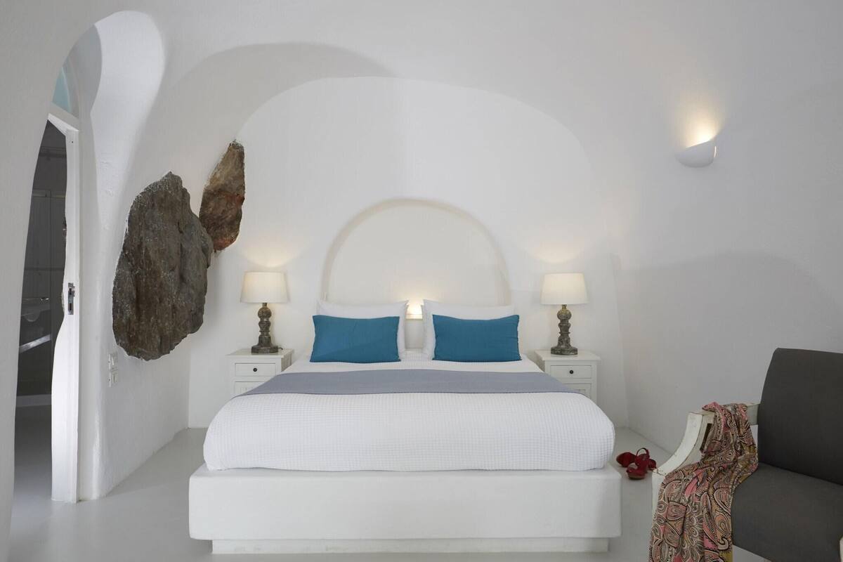 Quarto com cama branca, duas almofadas azuis, mesa de cabeceira branca com abajur, cadeira cinza e uma grande decoração de rocha na parede.