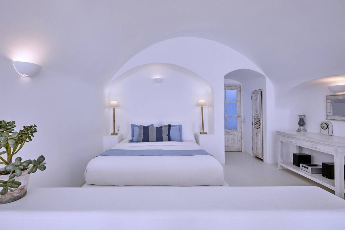 Quarto minimalista com uma cama grande, paredes brancas e uma porta em arco que dá para outro cômodo.