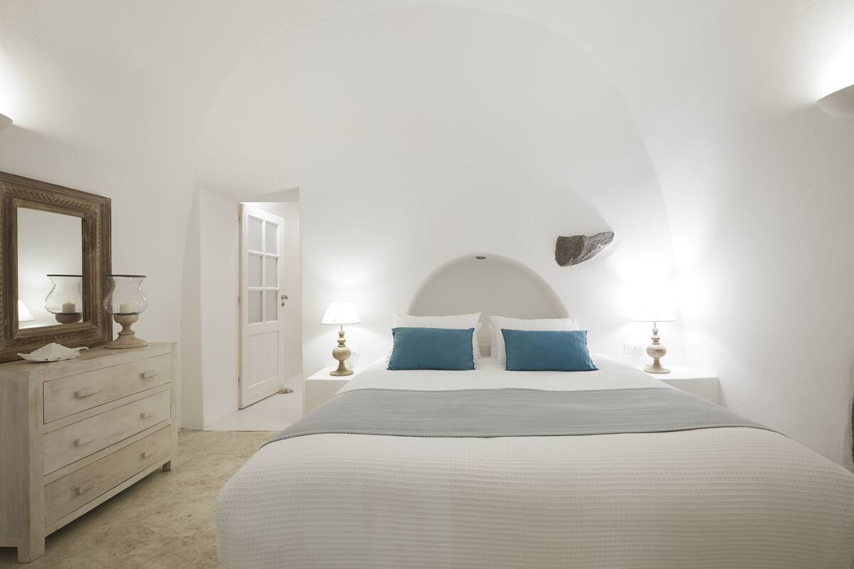 Quarto com cama, duas mesas de cabeceira com luminárias, cômoda, espelho e porta.