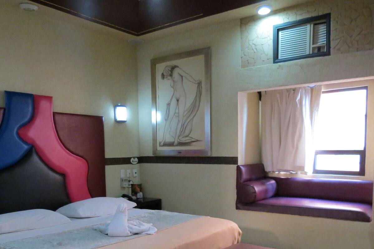 Quarto de hotel com uma cama grande, um banquinho, uma mesa de cabeceira, ar condicionado na parede e uma janela com vista.