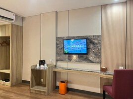 Uma área de recepção de hotel com televisão fixada numa parede de mármore, um guarda-roupa de madeira, um espaço de estar com uma cadeira vermelha e uma pequena lixeira.
