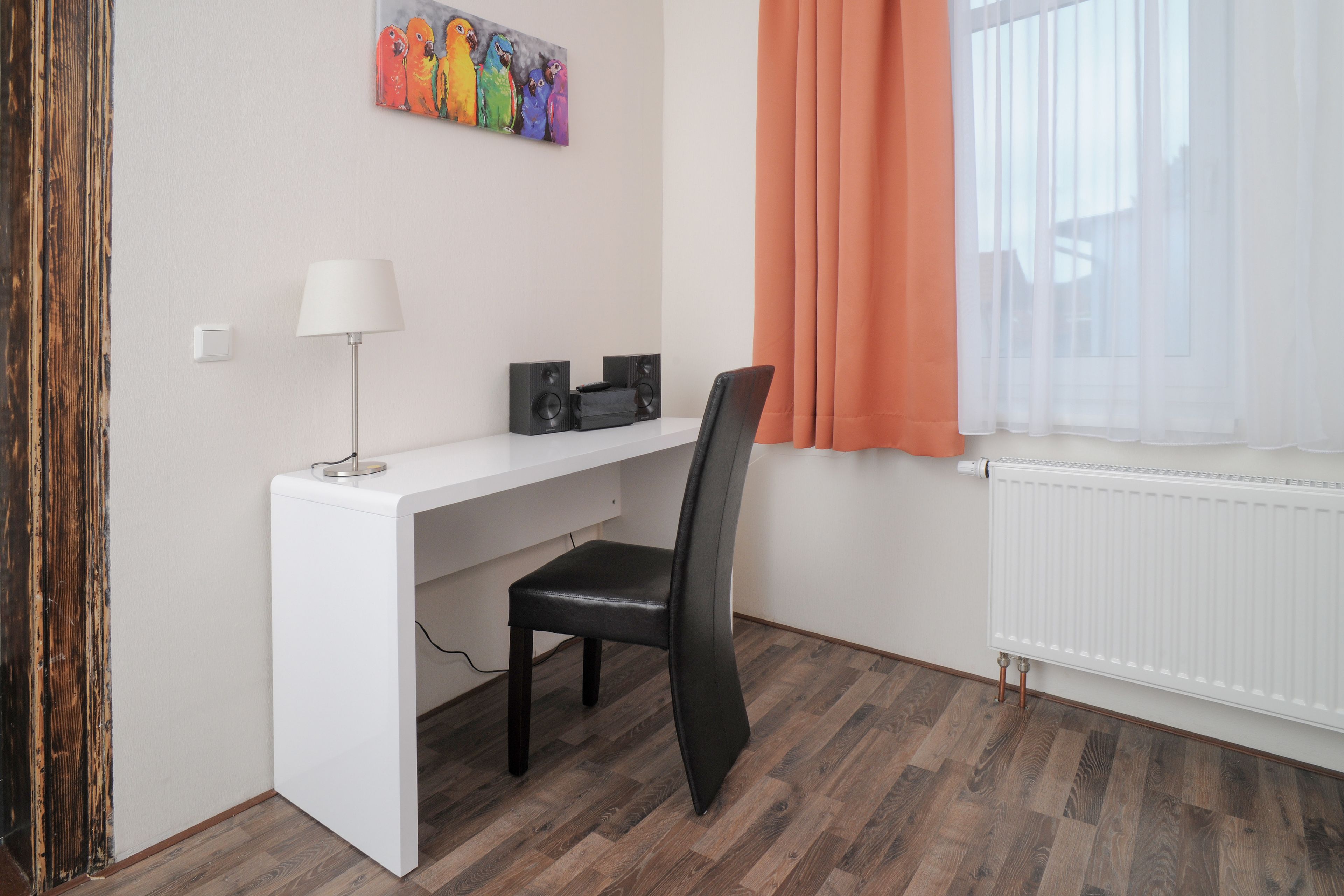 Quarto moderno com escrivaninha branca, cadeira preta e um quadro com pássaros coloridos na parede.