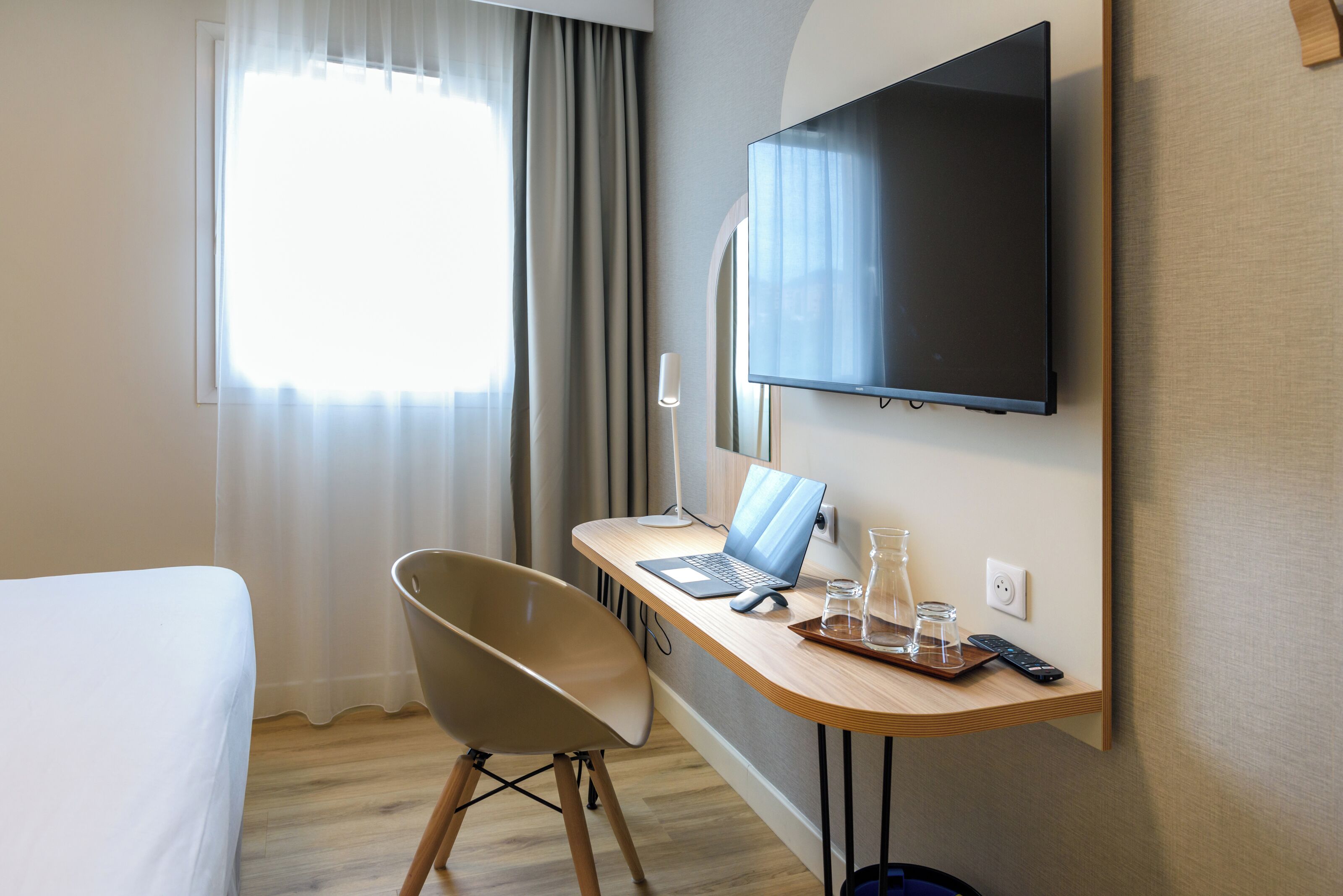 Quarto de hotel com uma cama, uma escrivaninha com um laptop, uma cadeira, uma TV fixada na parede e uma janela com cortinas.