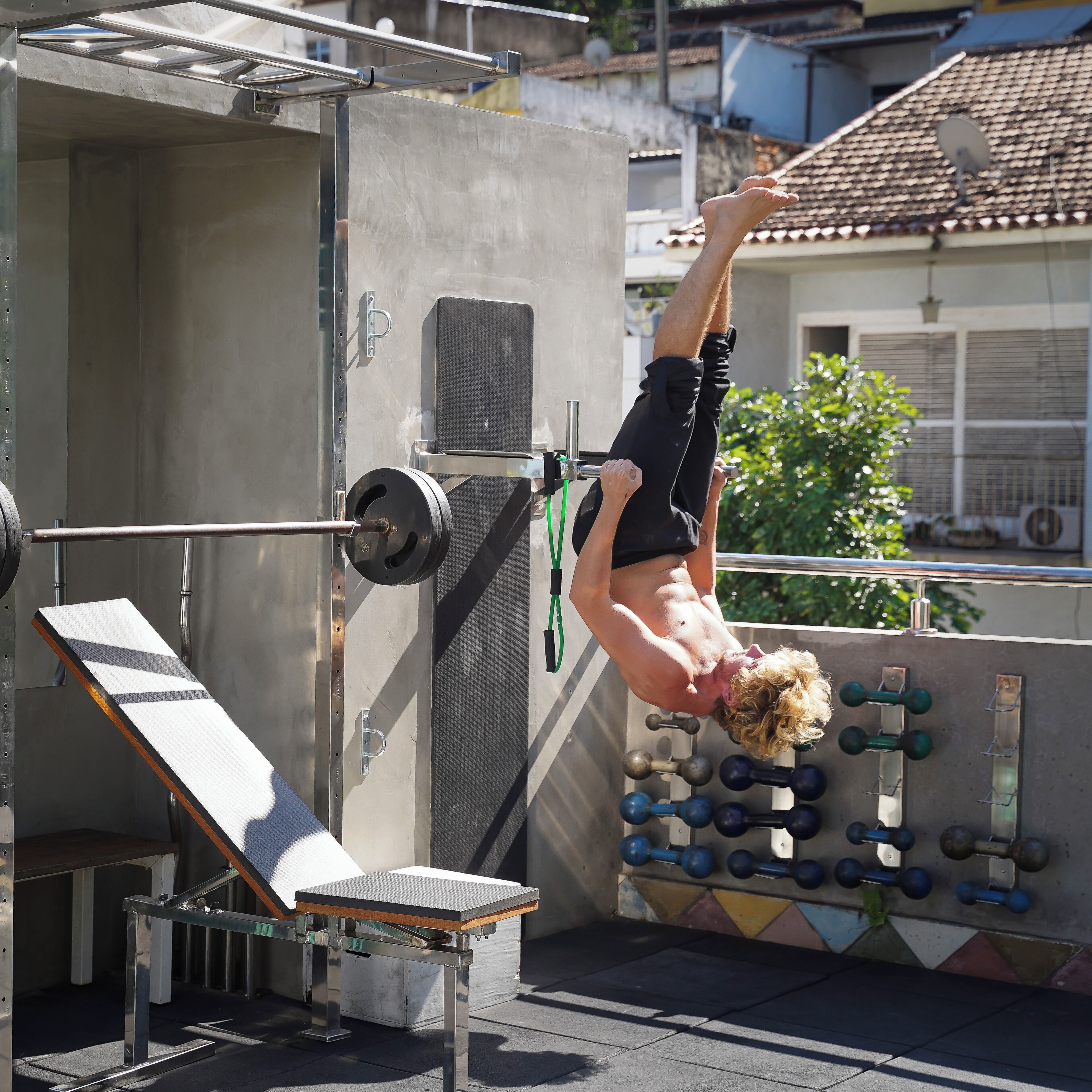 Uma pessoa fazendo uma ponte de mão em uma barra de metal, com um banco de musculação e halteres ao fundo.