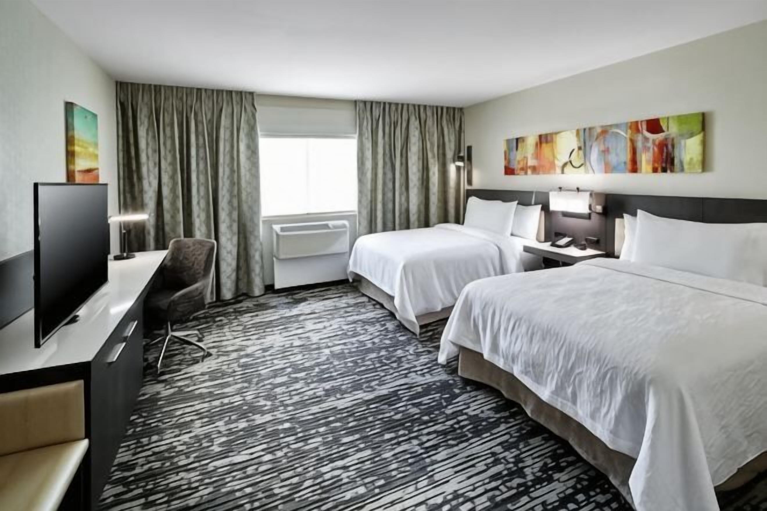 Quarto de hotel com duas camas, uma escrivaninha com televisão, uma cadeira e uma janela grande com cortinas.