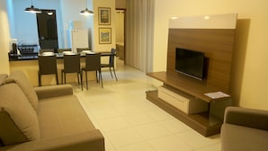 Uma sala de estar moderna com televisão de tela plana, um sofá, um espaço de refeições com cadeiras e uma cozinha ao fundo.