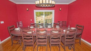 Uma sala de jantar com uma grande mesa de madeira posta para seis pessoas, rodeada por cadeiras combinando, um candeeiro e uma janela com vista.