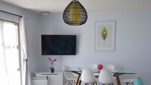 Uma sala de estar moderna com um sofá azul-turquesa, uma mesa de centro de vidro, uma área de jantar com mesa de vidro e cadeiras brancas, uma TV de tela plana na parede e um lustre.