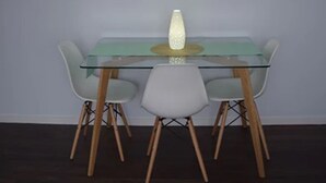Uma área de refeições com mesa de vidro e duas cadeiras, luminária na parede e um quadro emoldurado acima da mesa.
