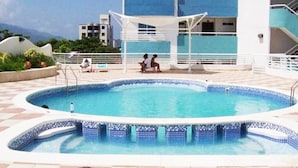 Uma piscina com borda azulejada, um toldo branco e um edifício com varandas ao fundo.