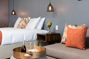 Quarto de hotel com cama, sofá, mesa de cabeceira, um vaso com flores e um tabuleiro com garrafa e copos.